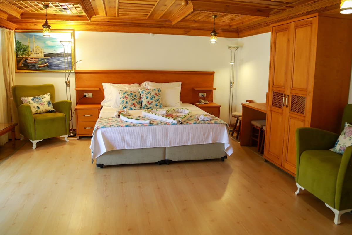 Quarto com cabeceira de madeira, cama com roupa de cama floral, duas poltronas verdes, guarda-roupa de madeira e um quadro na parede.