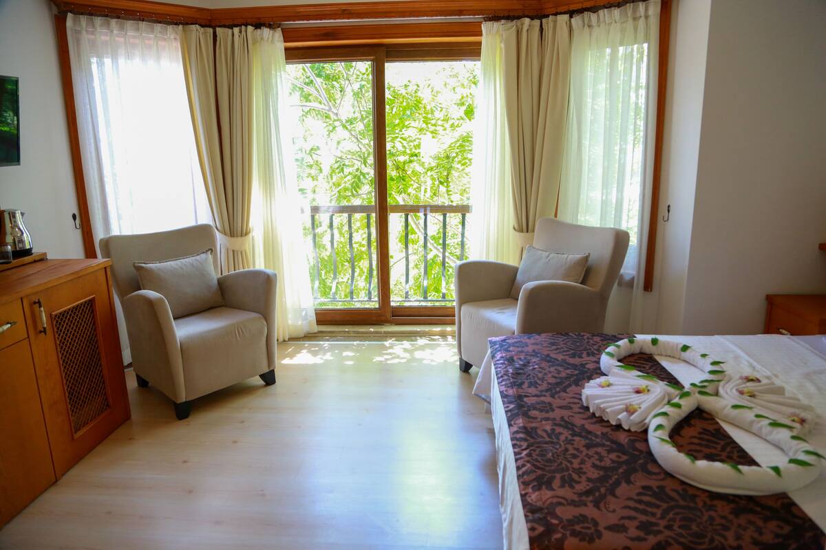 Quarto de hotel com varanda, duas poltronas, cama com colcha florida e armário de madeira.