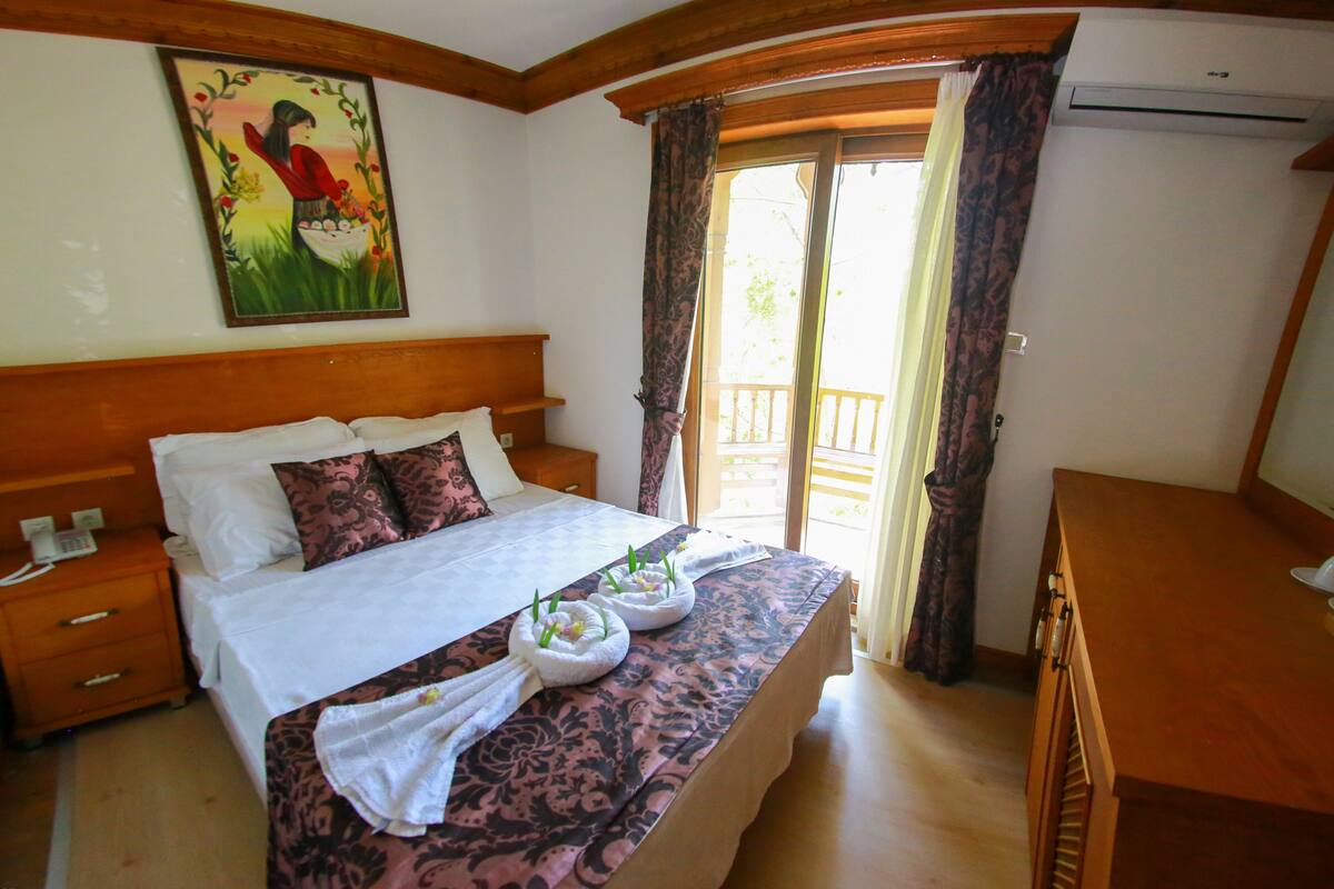 Quarto de hotel com uma cama grande, cabeceira de madeira, um quadro na parede e varanda com cortinas.