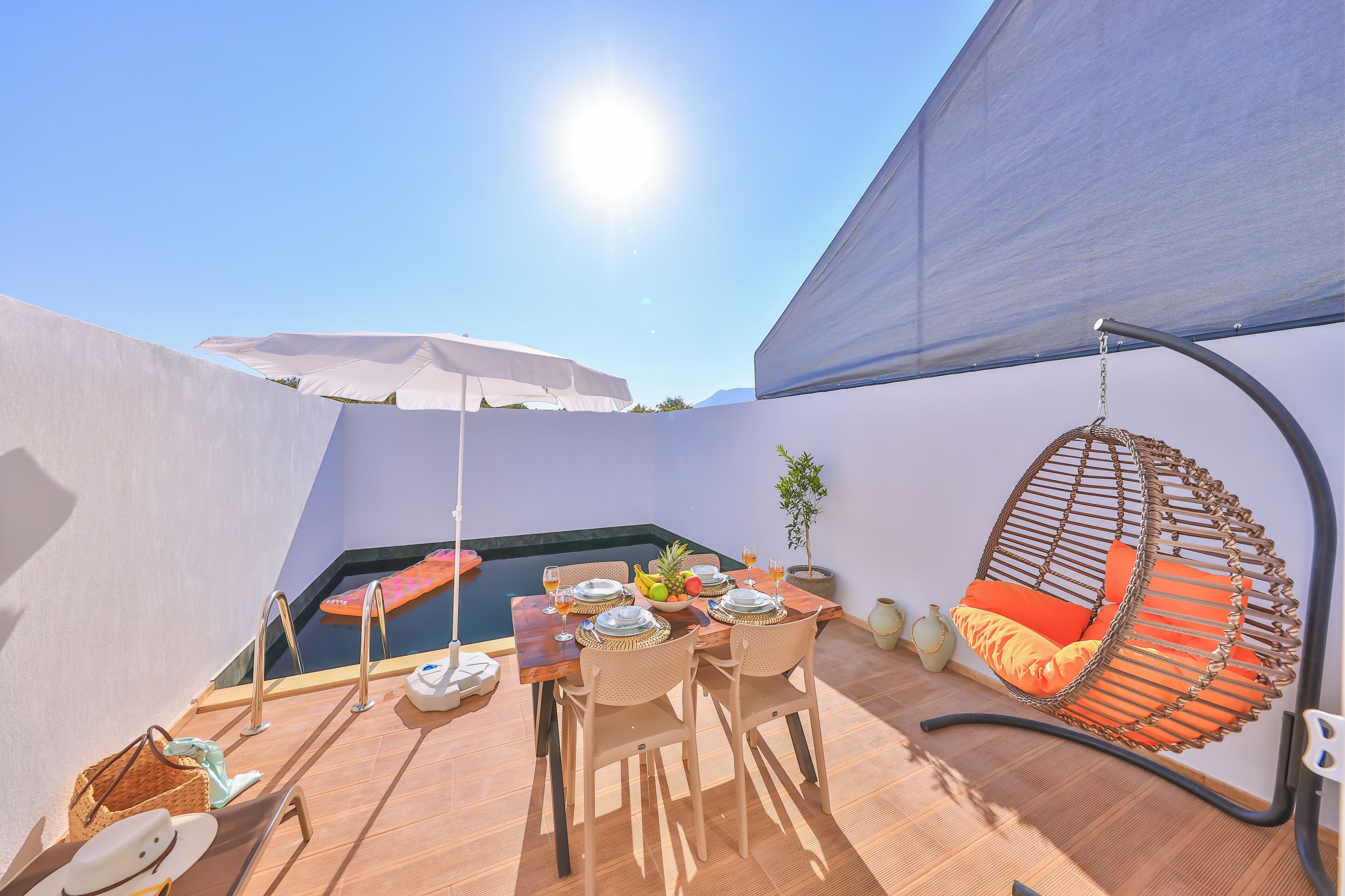 Uma área de refeições ao ar livre ensolarada com piscina, uma cadeira suspensa e guarda-sóis.