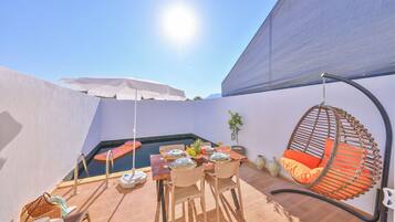 Uma área de refeições ao ar livre ensolarada com piscina, uma cadeira suspensa e guarda-sóis.