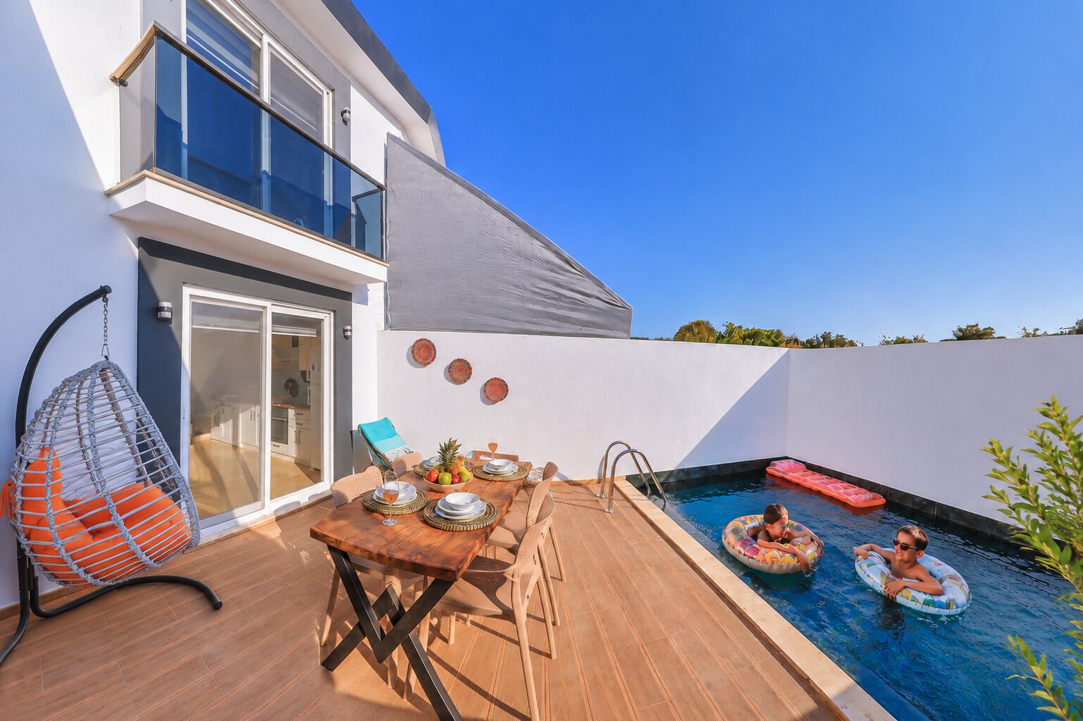 Uma área de piscina moderna ao ar livre com um deck de madeira, uma cadeira suspensa e uma mesa posta para refeições.