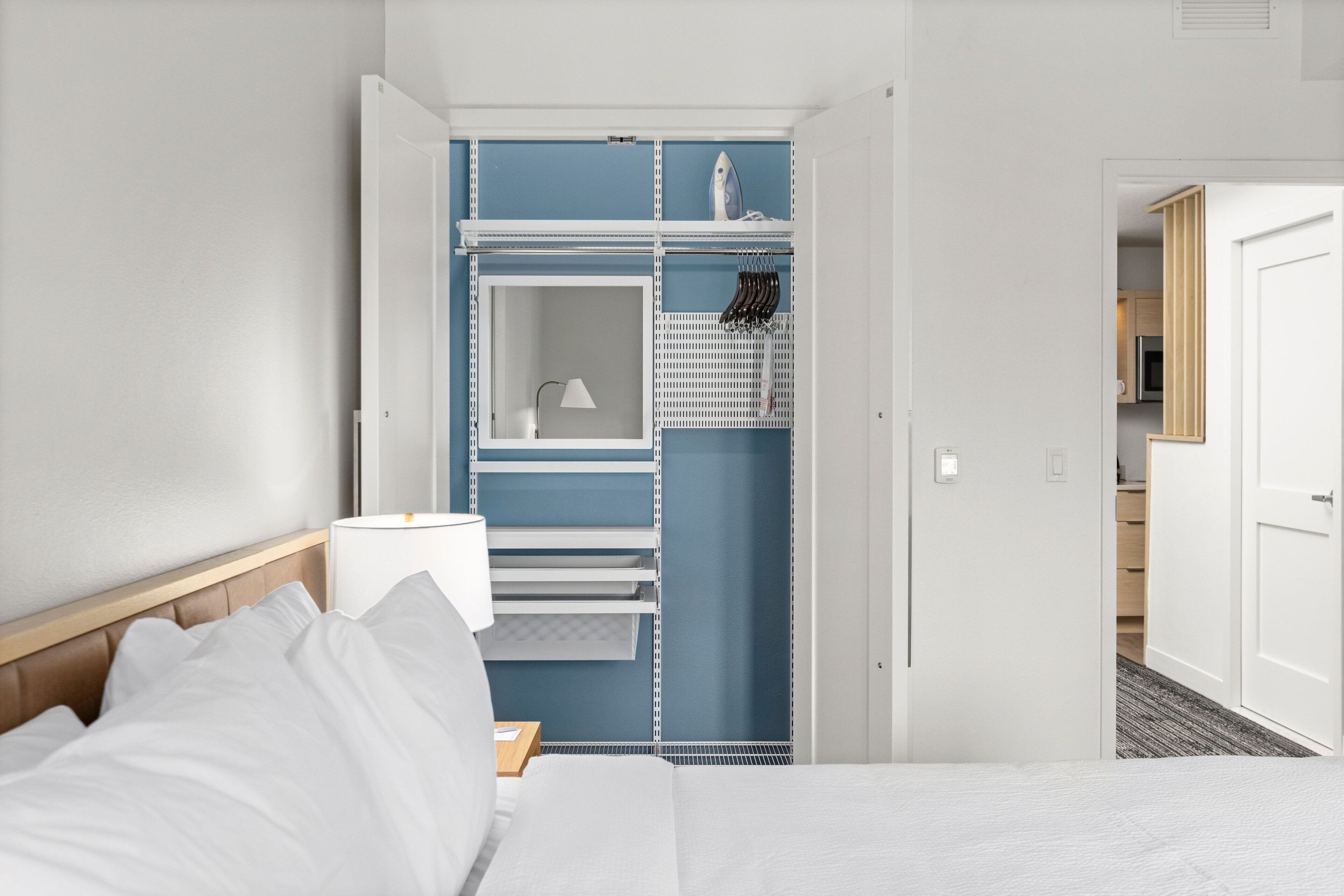 Quarto de hotel com uma cama, mesa de cabeceira, guarda-roupa com prateleiras e porta que dá para um banheiro.