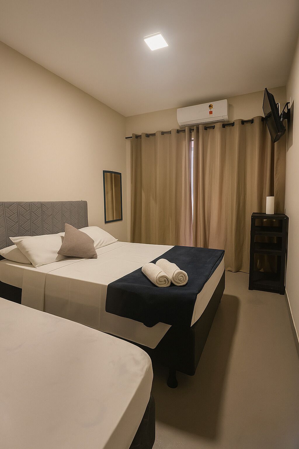 Quarto de hotel com duas camas, uma televisão de tela plana presa à parede, ventoinha no teto e janela com cortinas.