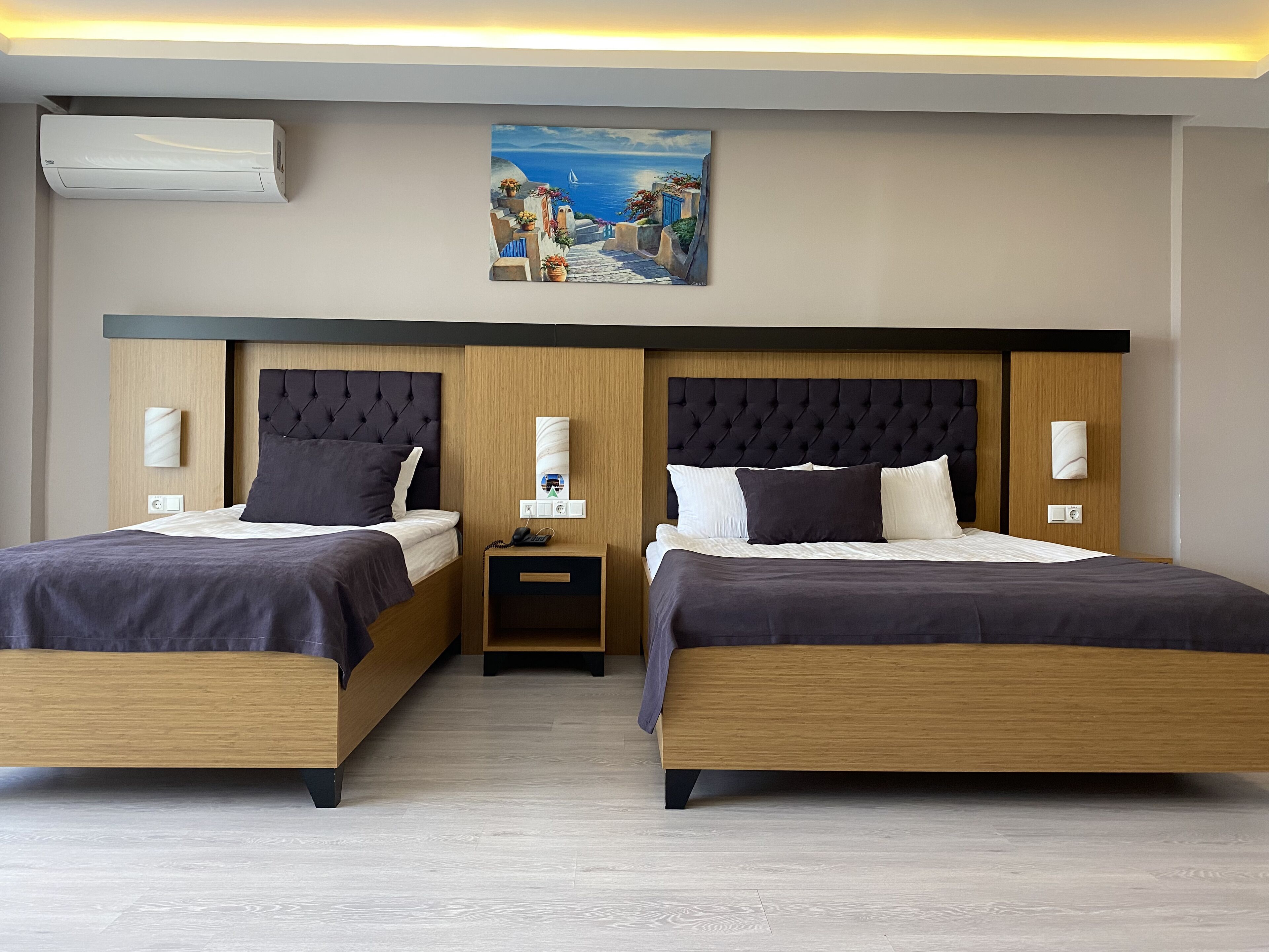 Quarto de hotel moderno com duas camas, cabeceiras de madeira, mesas de cabeceira e uma pintura de uma paisagem costeira.