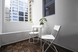 Quarto de canto com mesa e cadeira brancas, um vaso de planta e vista para prédios altos.