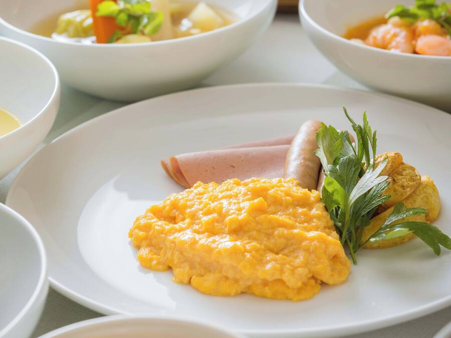 Mesa de café da manhã com ovos mexidos, presunto e acompanhamento de legumes no prato.