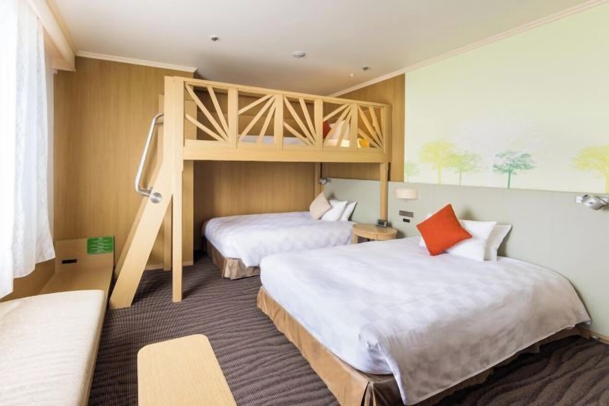 Quarto de hotel com duas camas, uma cama beliche, uma escrivaninha e uma mesinha de cabeceira.