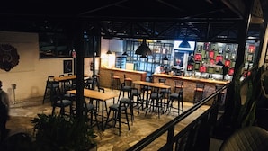 Um espaço de refeições interno com mesas e cadeiras, um balcão com uma pessoa atrás, e vista para a rua.