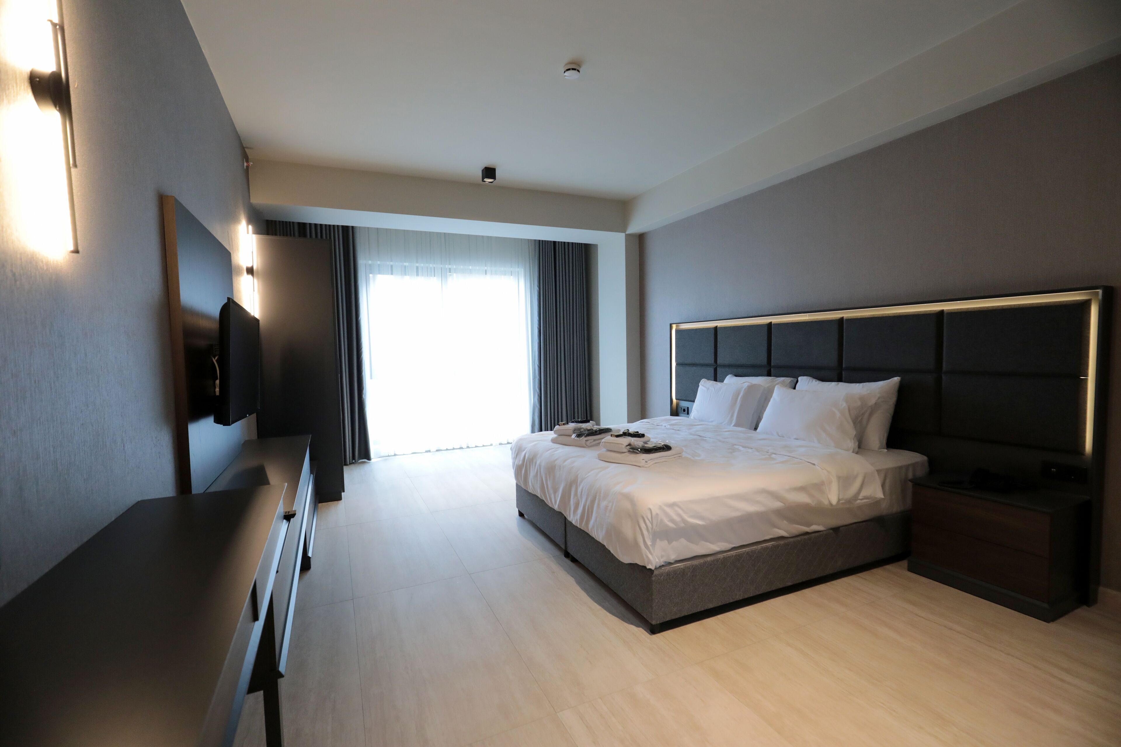 Quarto de hotel moderno com uma cama grande, televisão de tela plana, escrivaninha e uma grande janela com cortinas.