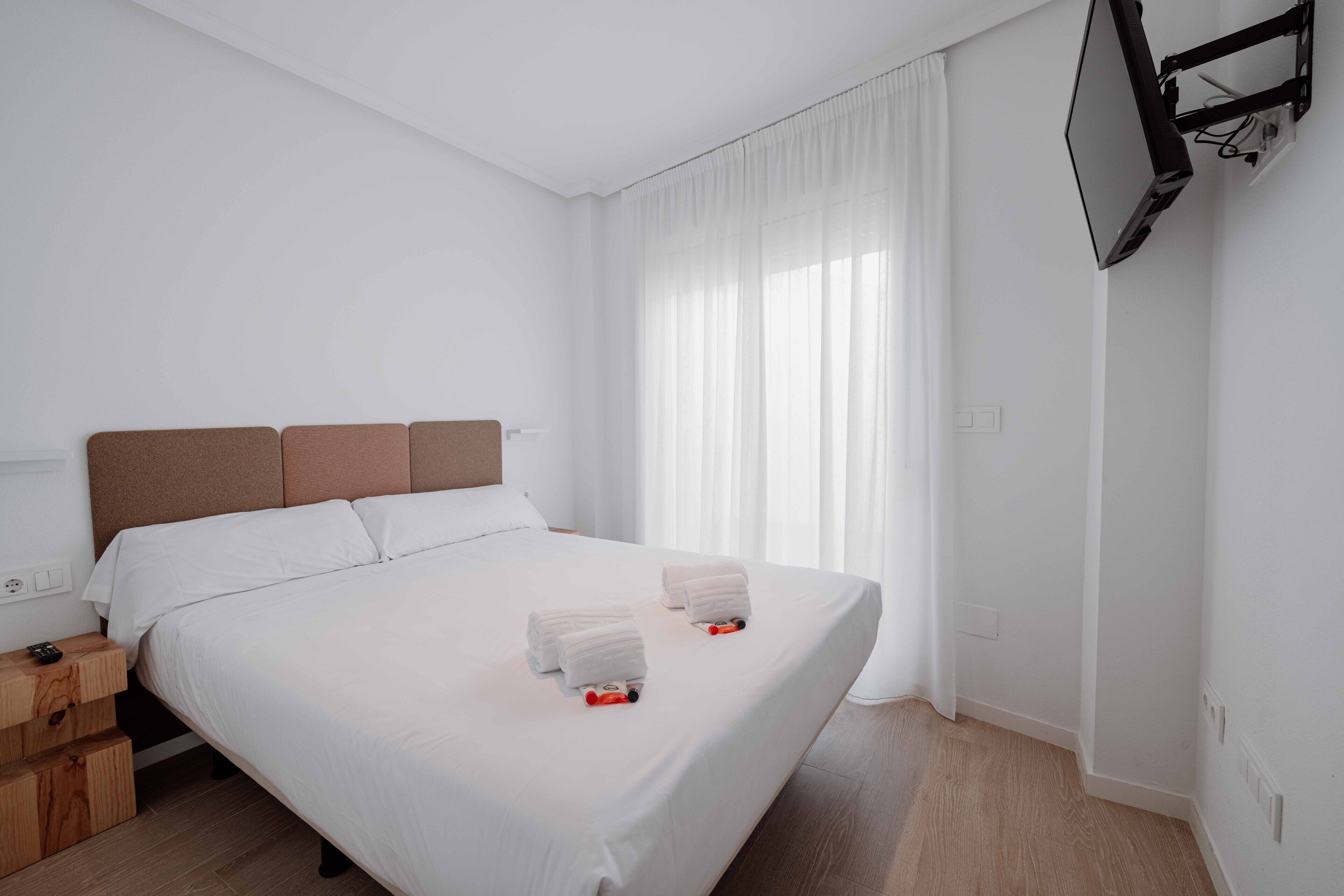 Cama bem arrumada com roupa de cama branca e duas toalhas, mesa de cabeceira de madeira com abajur, e televisão montada na parede.