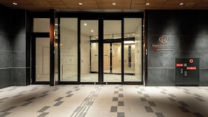 Entrada de hotel com design moderno, com porta de vidro, placa indicando o Hotel Daiwa Roynet e um elevador com sinalização de direção.