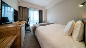 Quarto de hotel com uma cama, uma escrivaninha com computador, uma cadeira, uma televisão e uma janela com cortinas.