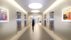 Corredor moderno de hotel, com elevadores, iluminação embutida e obras de arte nas paredes.