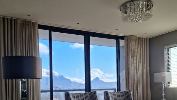 Quarto de hotel moderno com uma grande janela oferecendo vista da cidade, duas poltronas listradas, um sofá escuro e um candelabro.
