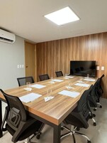 Sala de conferências com uma mesa grande, cadeiras, TV de tela plana e luminária no teto.