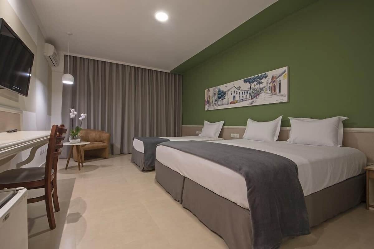 Quarto de hotel com cama, escrivaninha, cadeira, mesinha com uma planta e um quadro na parede.
