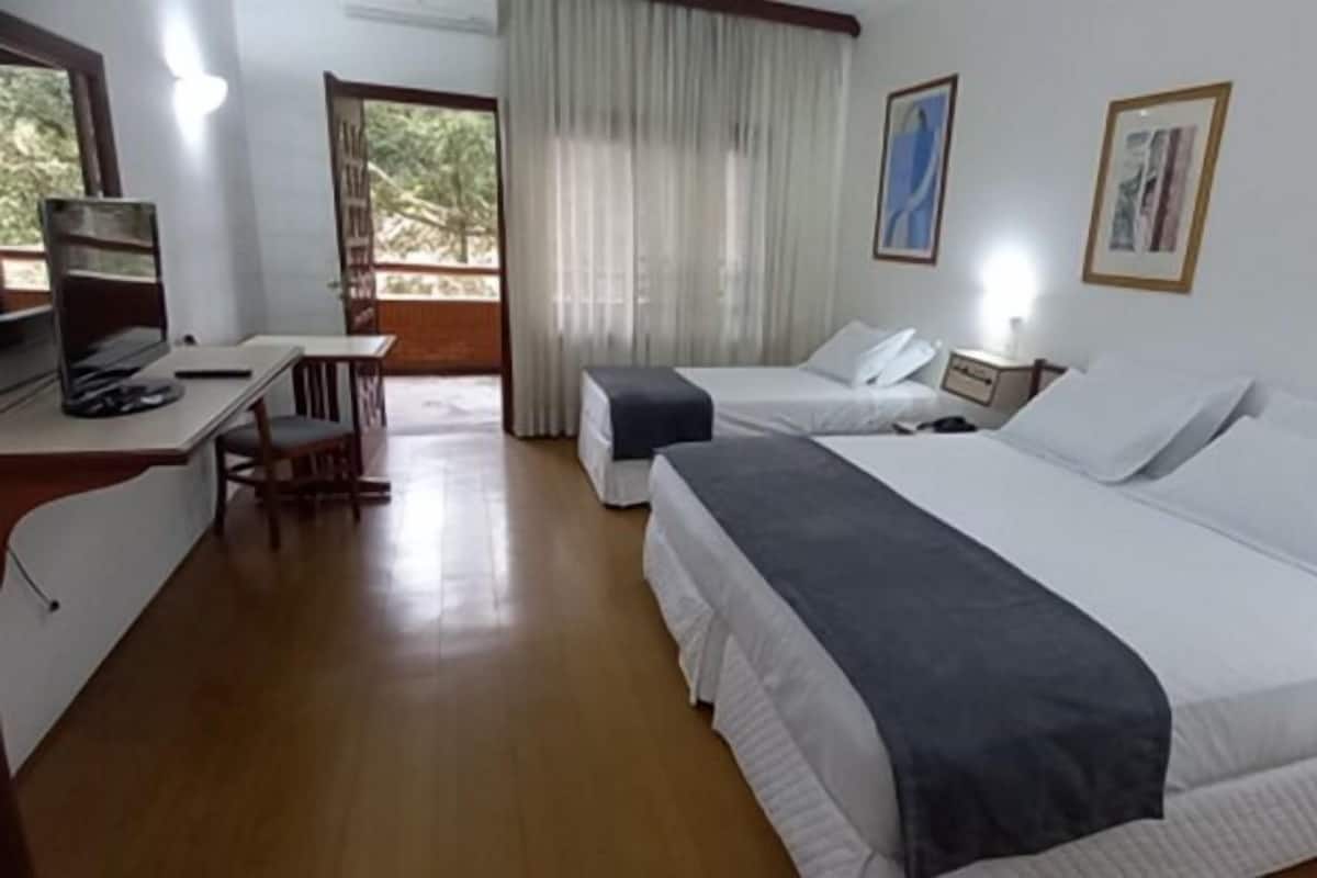 Quarto de hotel com duas camas, uma escrivaninha com cadeira, televisão e janela com cortinas.