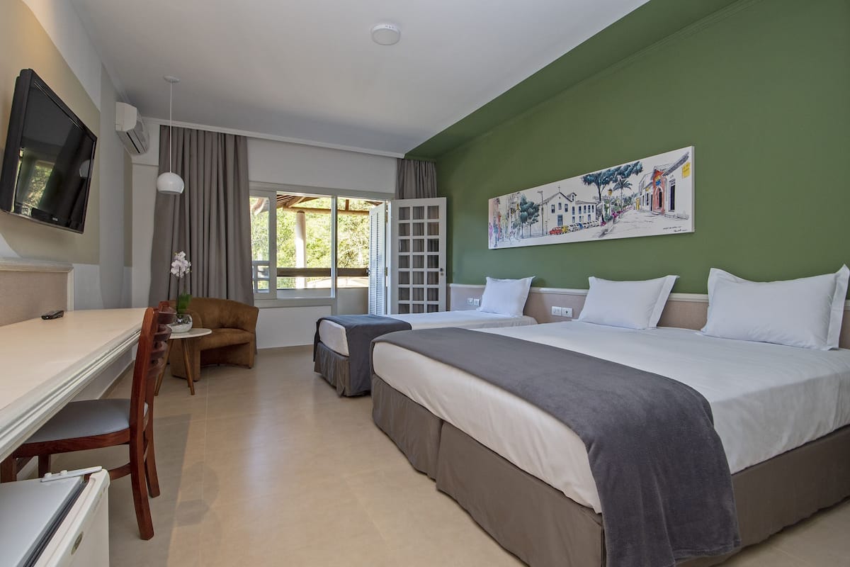 Quarto de hotel com cama, escrivaninha, cadeira, televisão e janela com cortinas.