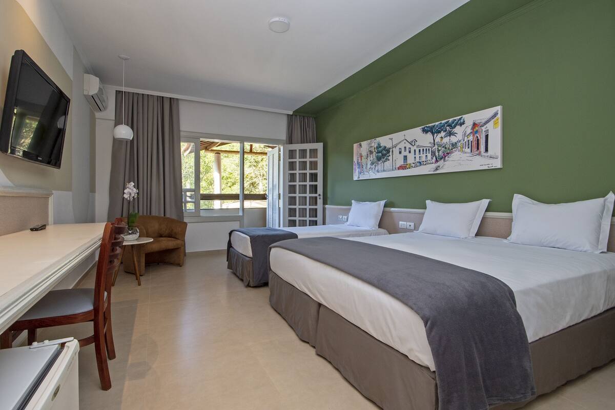 Quarto de hotel com cama, escrivaninha, cadeira, televisão e janela com cortinas.