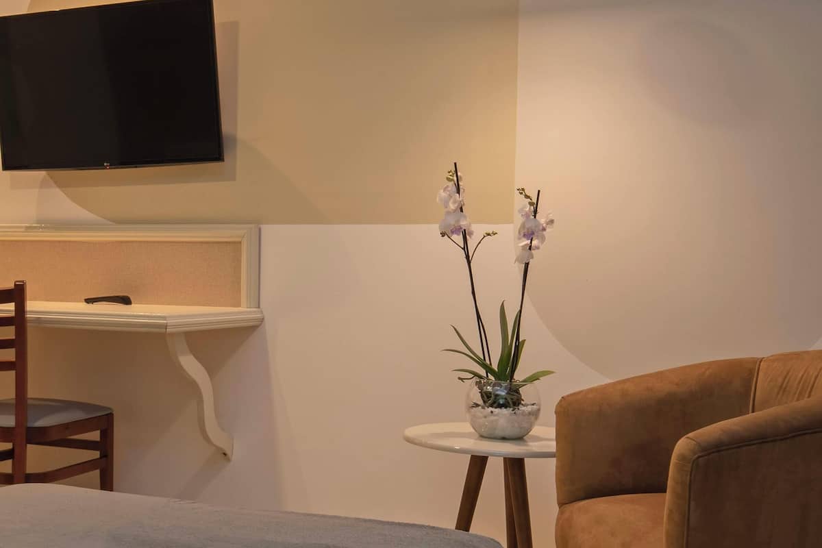 Quarto de hotel com uma cama, uma escrivaninha com cadeira, uma poltrona marrom, uma mesinha com uma orquídea em vaso e uma televisão presa à parede.