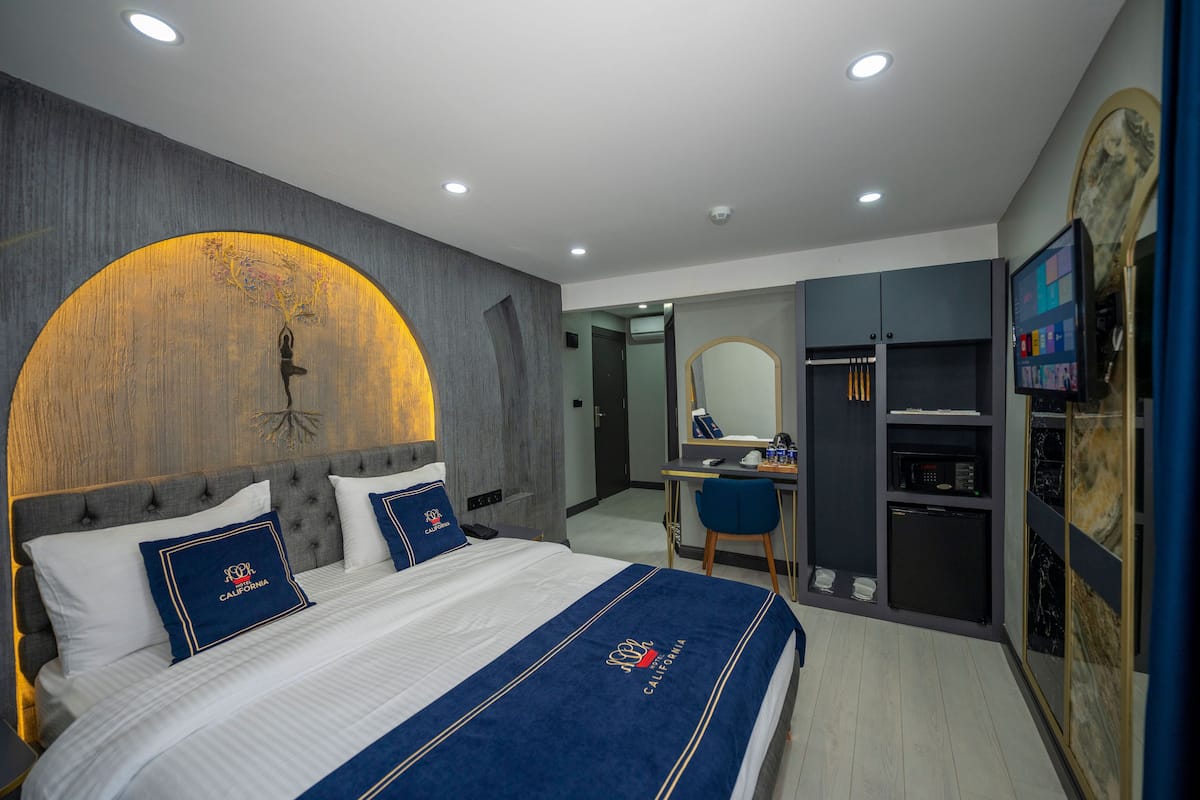 Quarto de hotel com uma cama grande, uma escrivaninha, uma cadeira, um espelho e uma televisão.