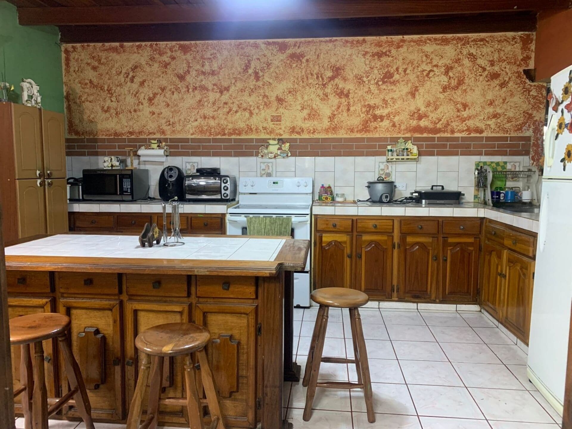 Uma cozinha com armários de madeira, painel de azulejos, uma ilha central com bancos e diversos eletrodomésticos.