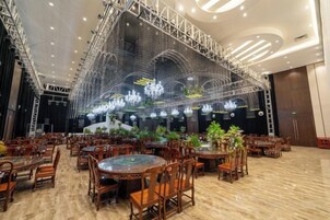 Uma ampla sala de banquetes com várias mesas redondas e cadeiras, teto alto com estrutura metálica decorativa e um lustre.