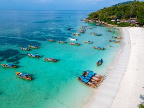 Uma praia com vários barcos ancorados perto da costa, cercada por vegetação exuberante e um mar azul cristalino.
