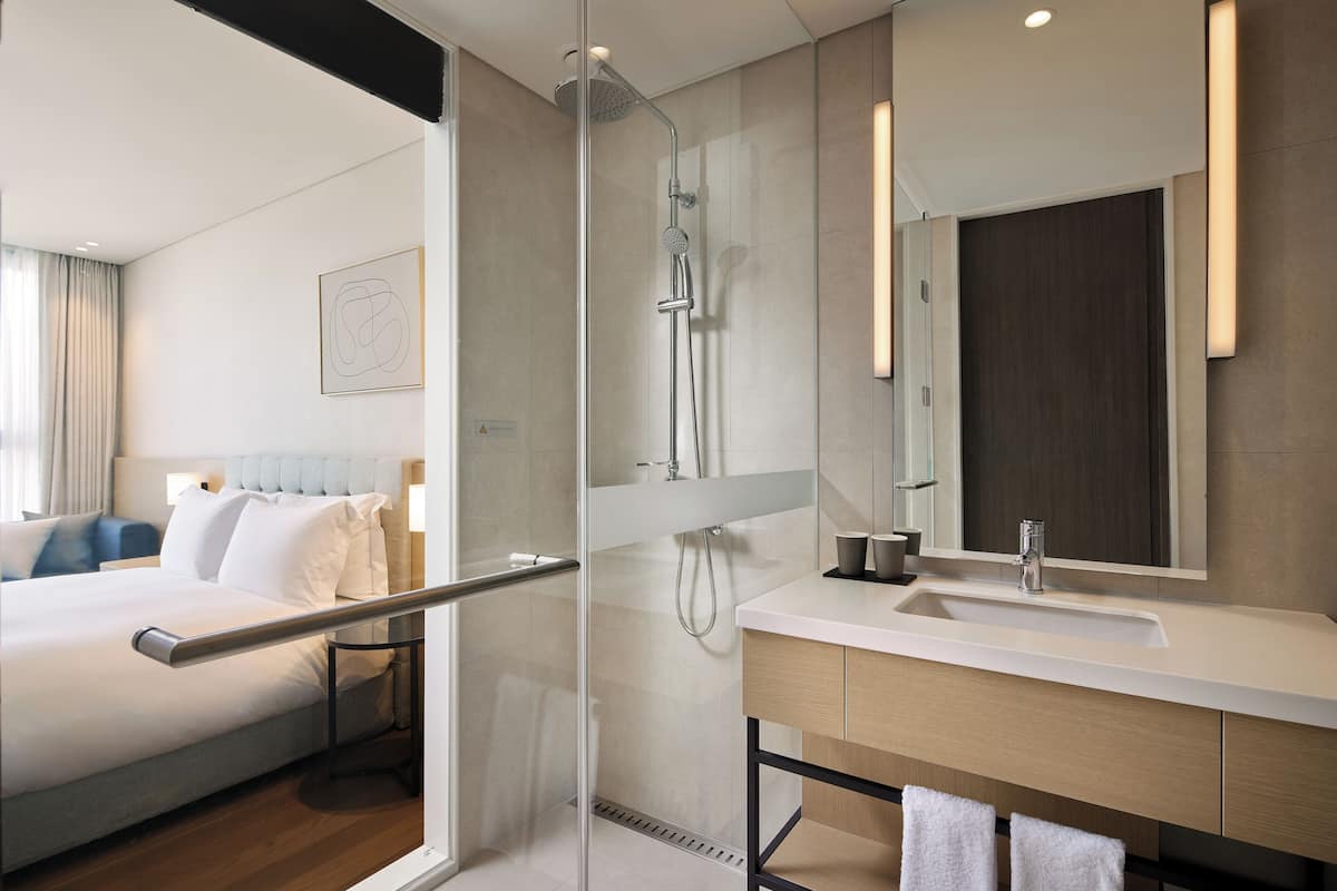 Quarto de hotel moderno com chuveiro em box de vidro, uma cama grande com roupa de cama branca, cabeceira de madeira e um espelho grande sobre a pia.