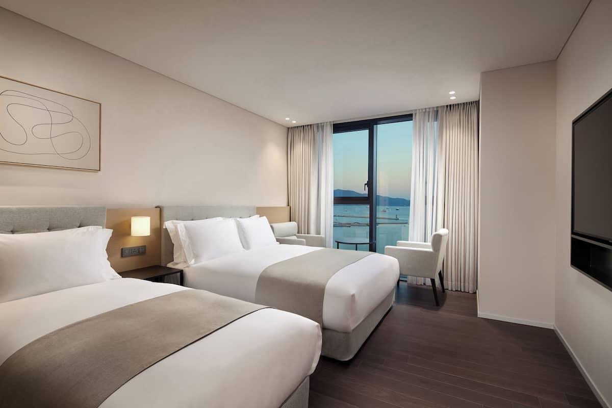 Quarto de hotel com duas camas, uma escrivaninha, uma cadeira, televisão e uma janela grande com vista para o mar.