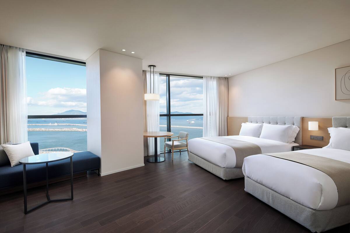 Quarto de hotel com duas camas, um sofá, uma mesa e vista para o mar.