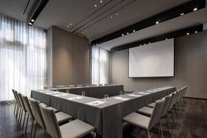 Uma sala de conferência com uma mesa comprida, cadeiras, uma tela de projetor e uma janela grande com cortinas.