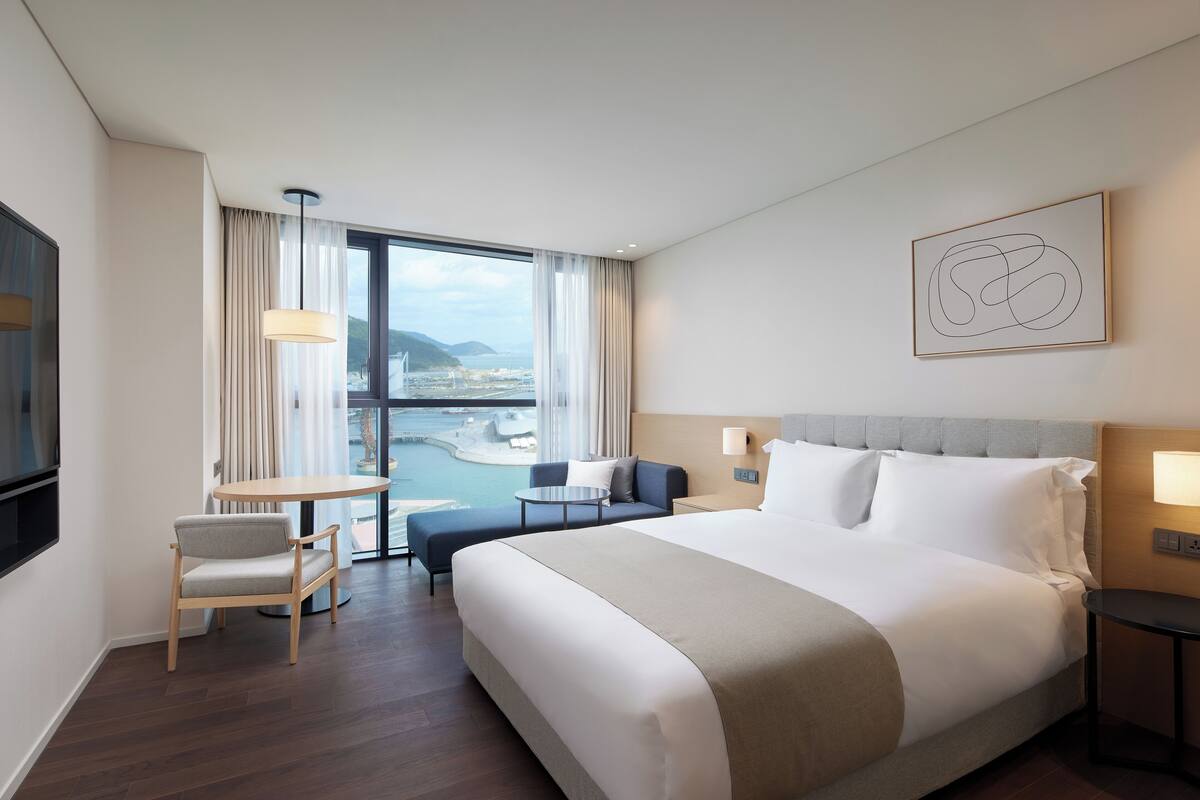 Quarto de hotel com uma cama grande, área de estar com uma mesa e vista para um corpo d'água e paisagem ao longe.