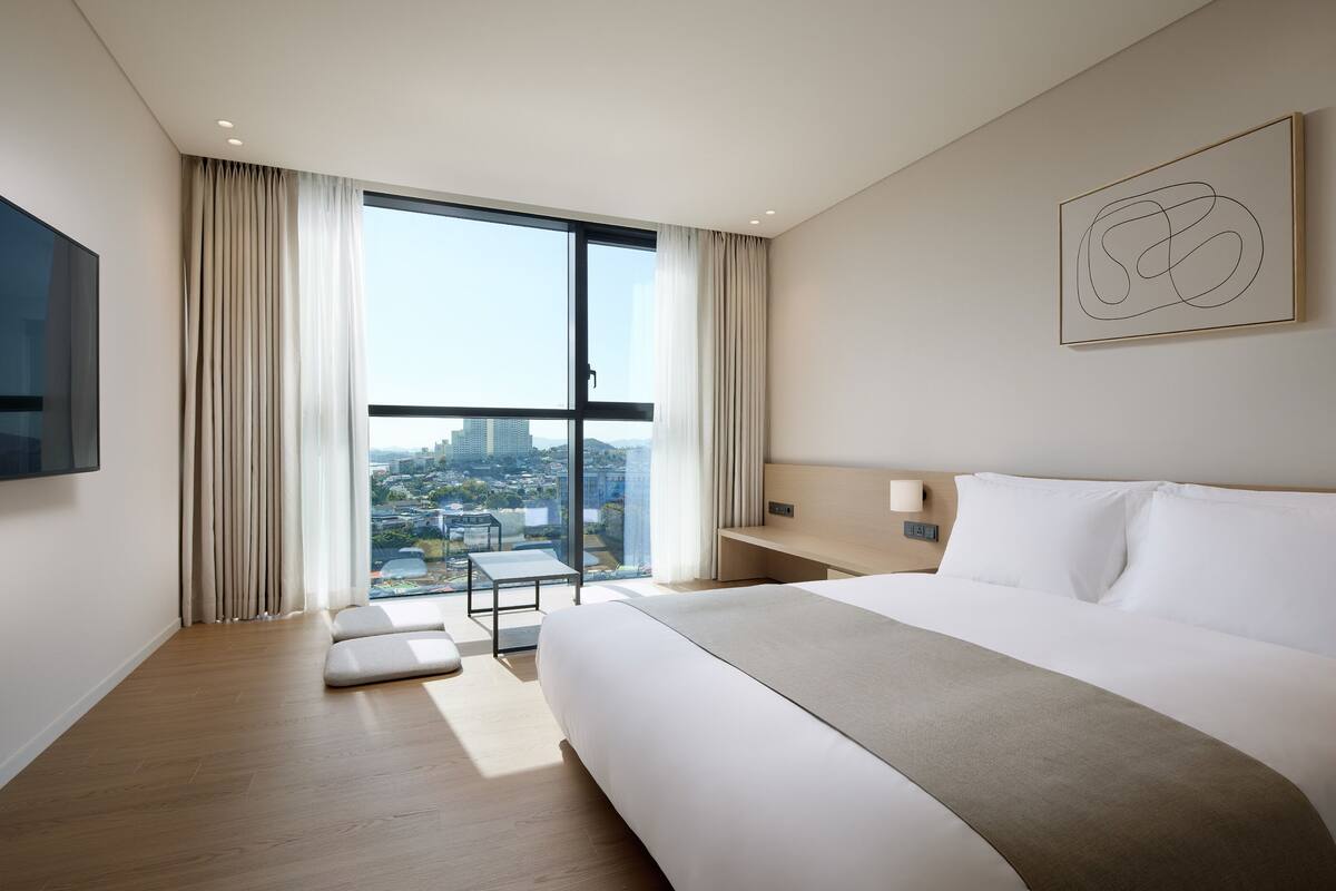 Quarto de hotel moderno com uma cama grande, uma escrivaninha e vista para a cidade através de uma janela do chão ao teto.