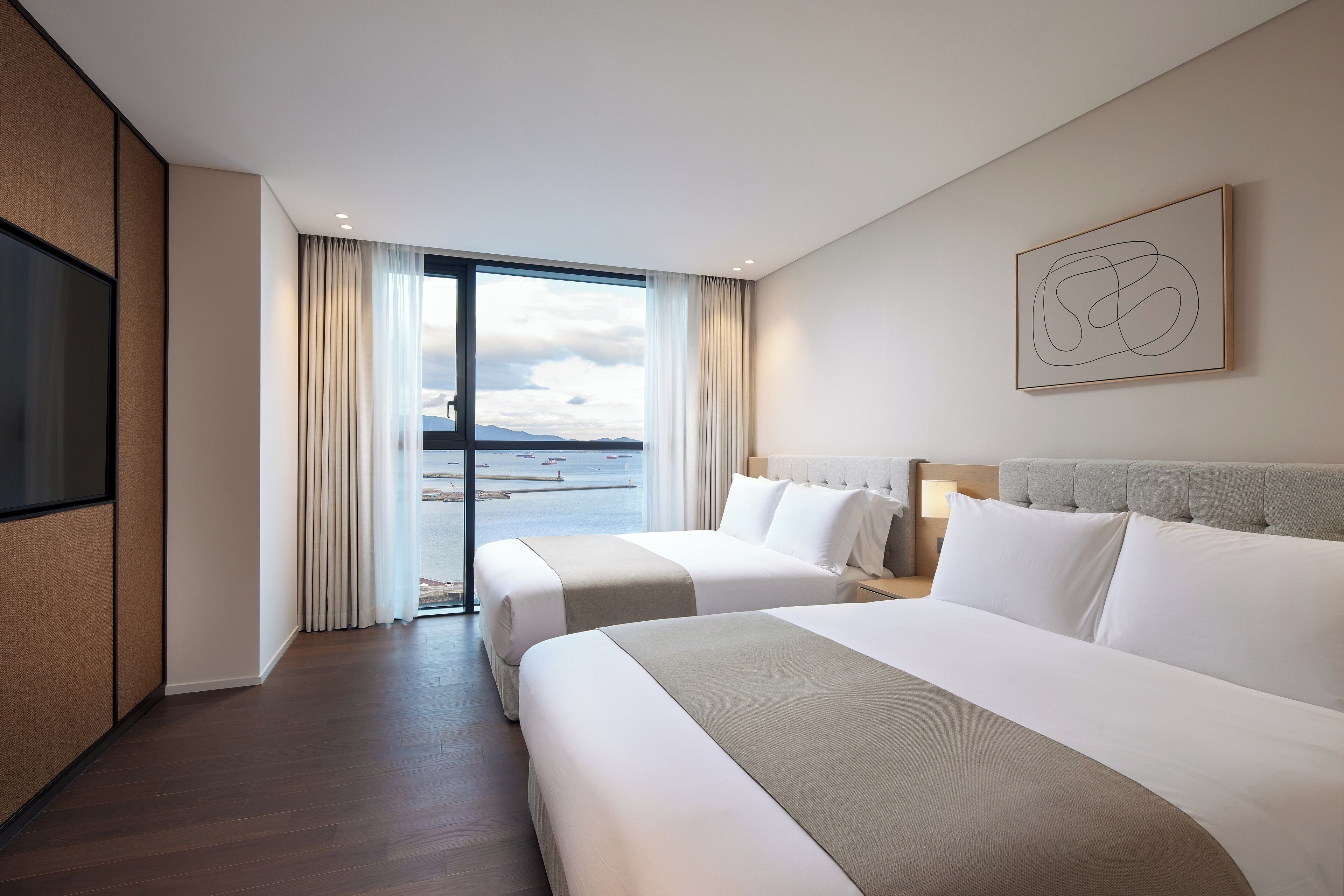 Quarto de hotel com duas camas, uma janela ampla com cortinas, uma televisão de tela plana e uma obra de arte abstrata na parede.