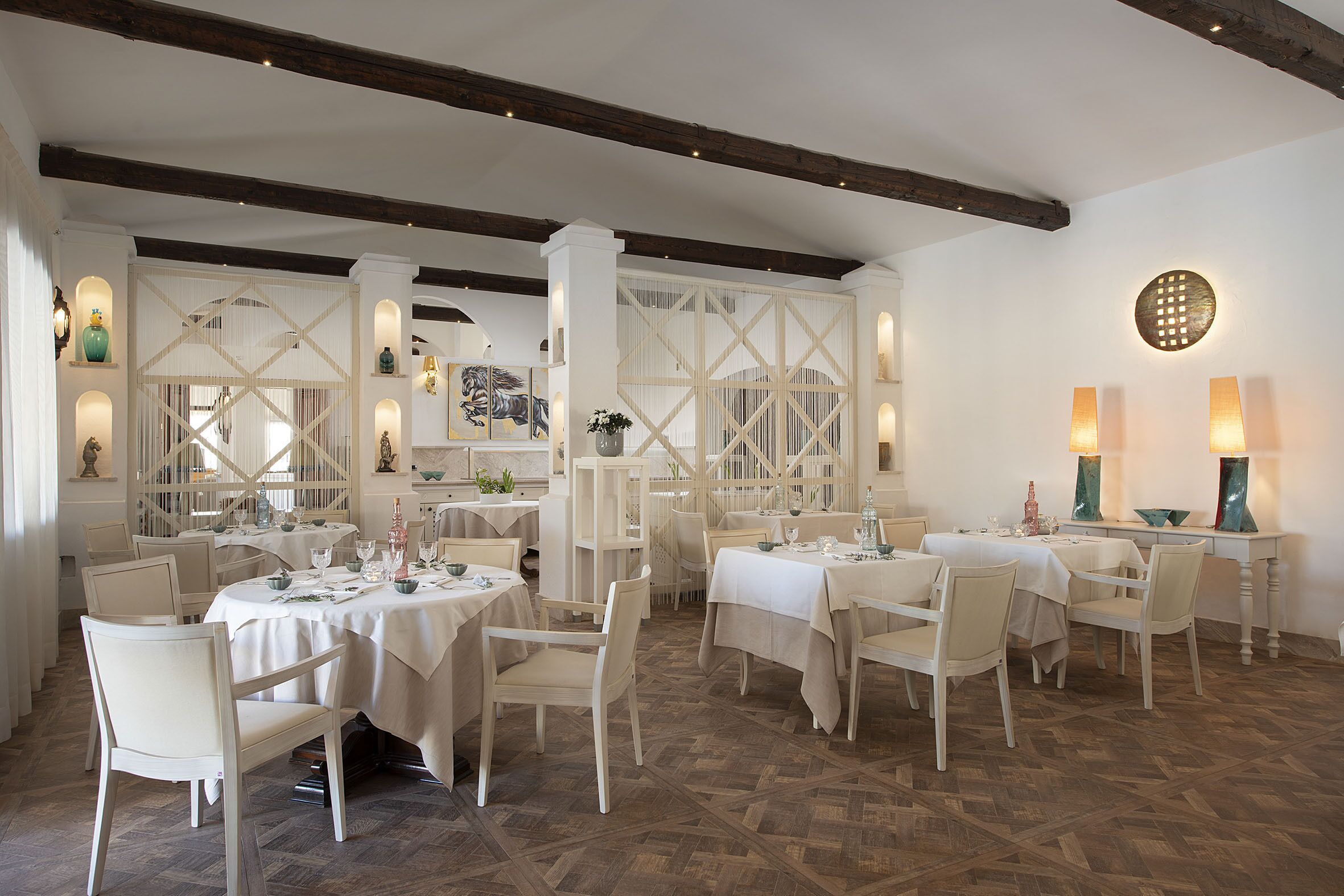 Uma sala de jantar com mesas e cadeiras brancas, vigas de madeira no teto e iluminação fixada na parede.