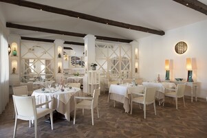 Uma sala de jantar com mesas e cadeiras brancas, vigas de madeira no teto e iluminação fixada na parede.