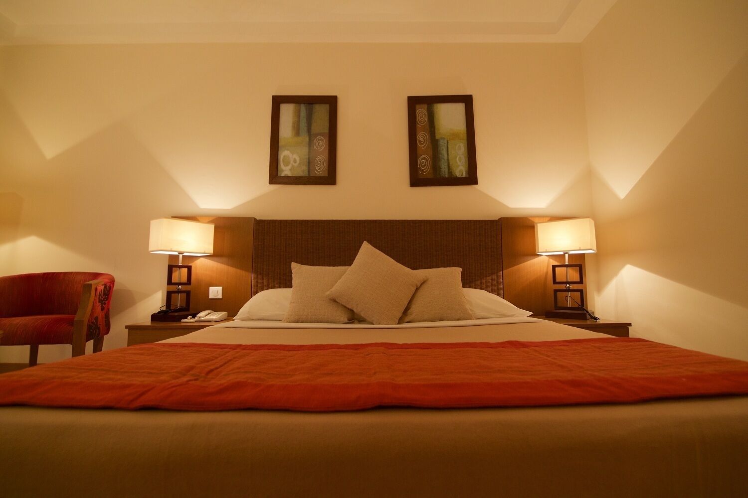 Quarto de hotel com uma cama grande, duas luminárias de cabeceira, uma cadeira e duas obras de arte emolduradas na parede.