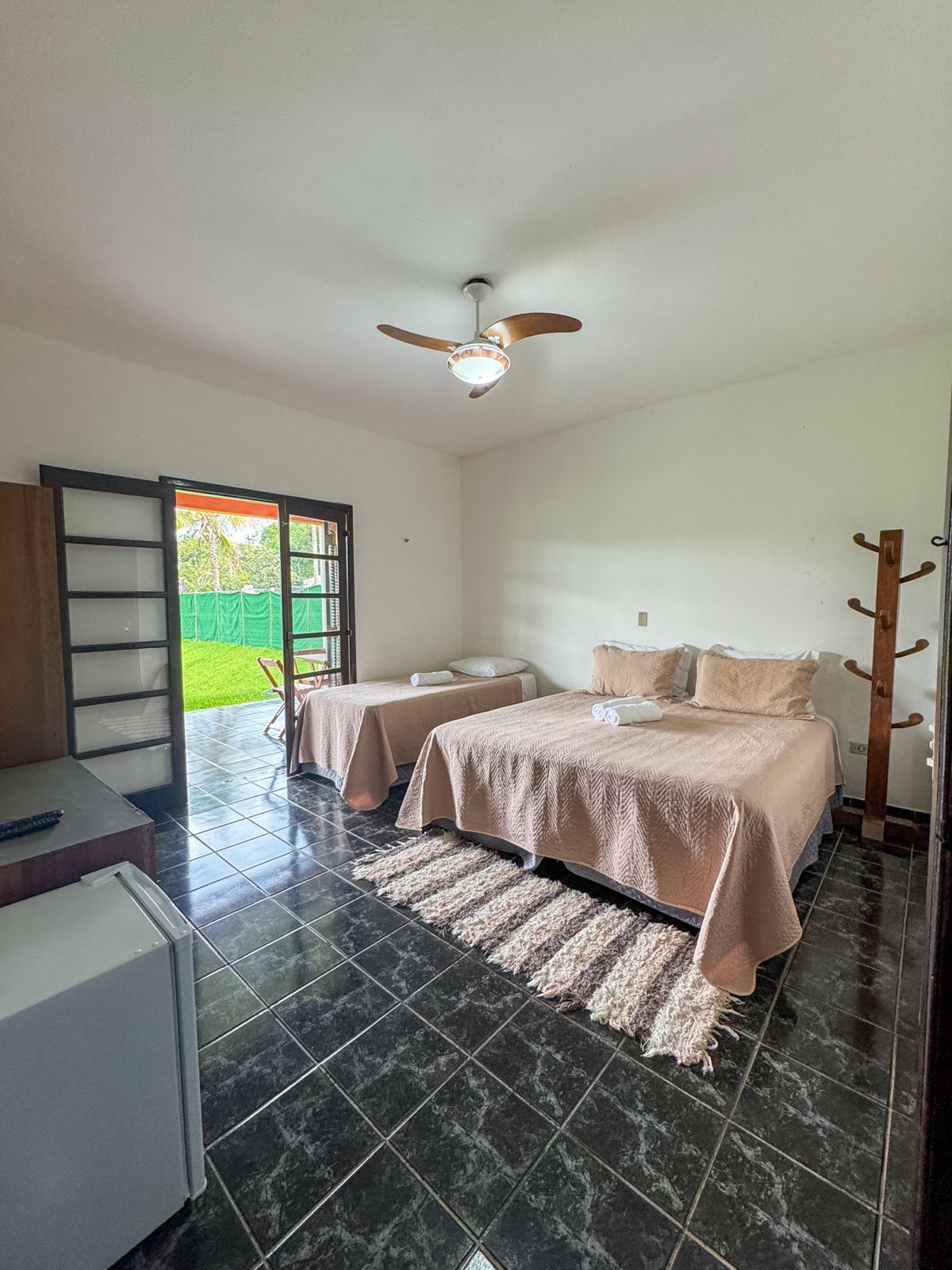 Quarto de hotel com duas camas, um ventilador de teto, uma mesinha, um cabideiro e vista para área externa verde.