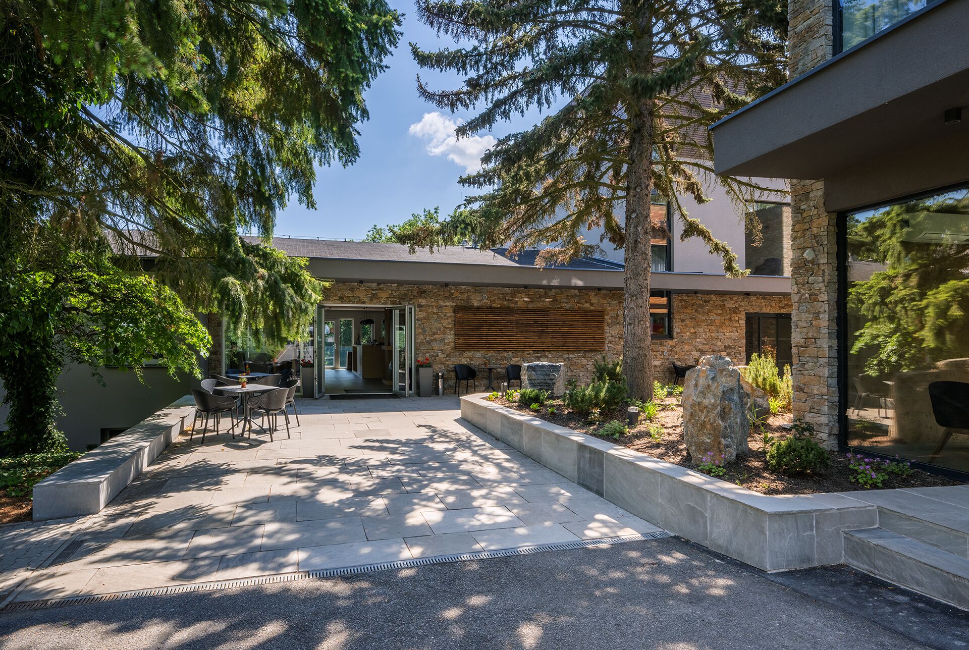 Uma casa moderna com fachada de pedra, um pátio com mobiliário de exterior e vista para as árvores ao redor.