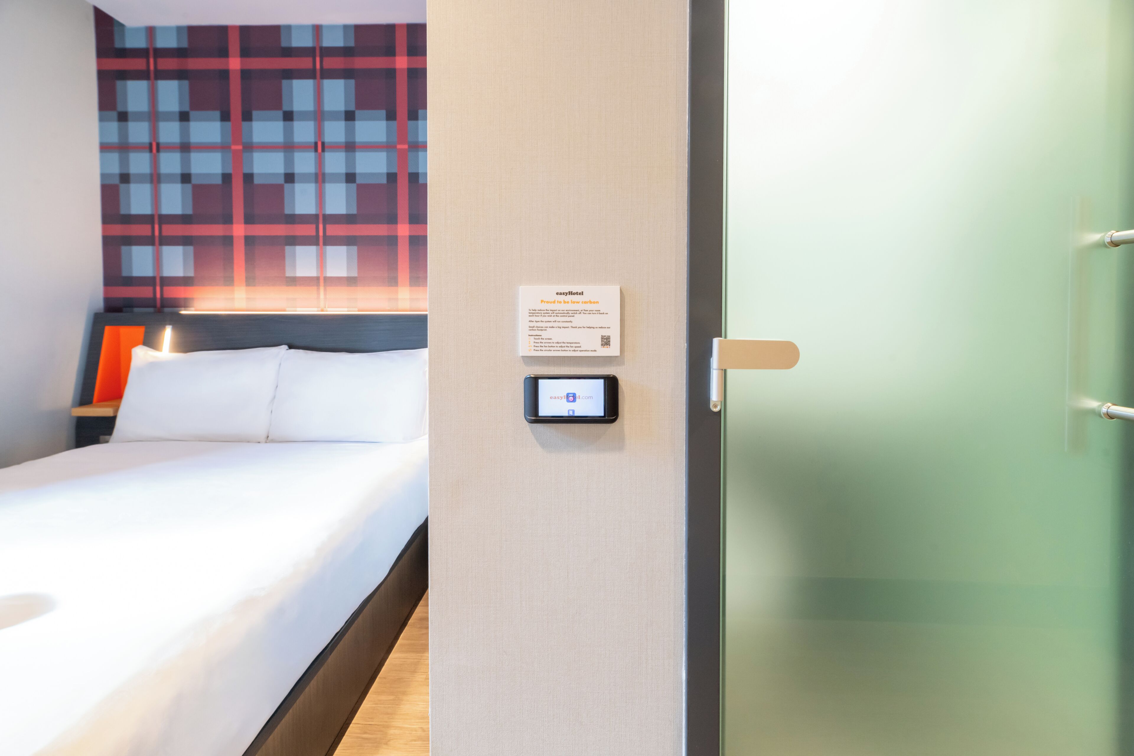 Quarto de hotel com cama, painel de controle fixado na parede e uma porta.