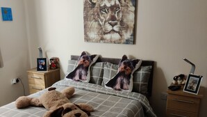 Quarto com cama, almofadas, um ursinho de pelúcia, mesa de cabeceira, abajur e um quadro na parede.