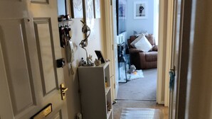 Um corredor com uma porta à esquerda, um armário branco com diversos objetos, e uma sala de estar ao fundo com um sofá e uma televisão.