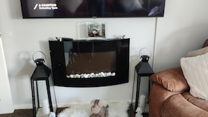 Uma sala de estar com televisão de tela plana na parede, lareira com um quadro emoldurado acima, duas lanternas pretas, um sofá marrom e um tapete branco e fofo.