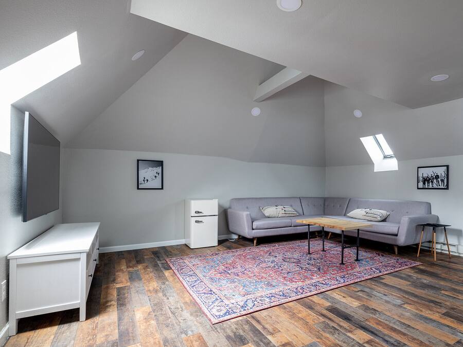 Quarto estilo loft com sofá cinza, escrivaninha branca pequena e tapete colorido.