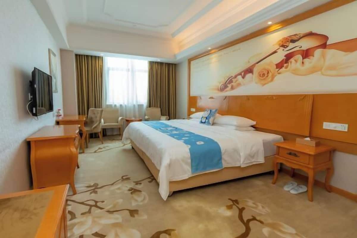 Quarto de hotel com uma cama grande, uma escrivaninha, uma cadeira, uma televisão e um quadro na parede.