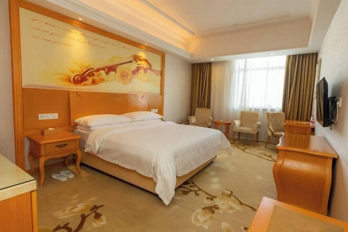 Quarto de hotel com uma cama grande, uma escrivaninha, uma cadeira, uma televisão e um objeto decorativo na parede.