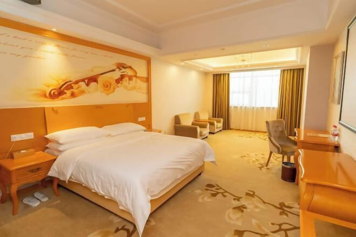 Quarto de hotel com uma cama grande, uma escrivaninha, uma cadeira e um sofá. Há um quadro na parede e uma janela com cortinas.