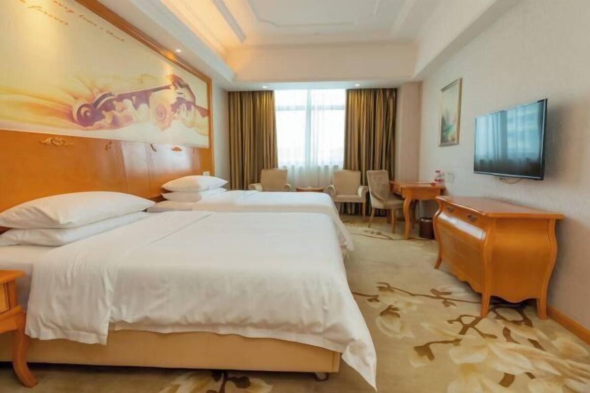 Quarto de hotel com duas camas, uma escrivaninha, uma cadeira, uma televisão e um quadro na parede.